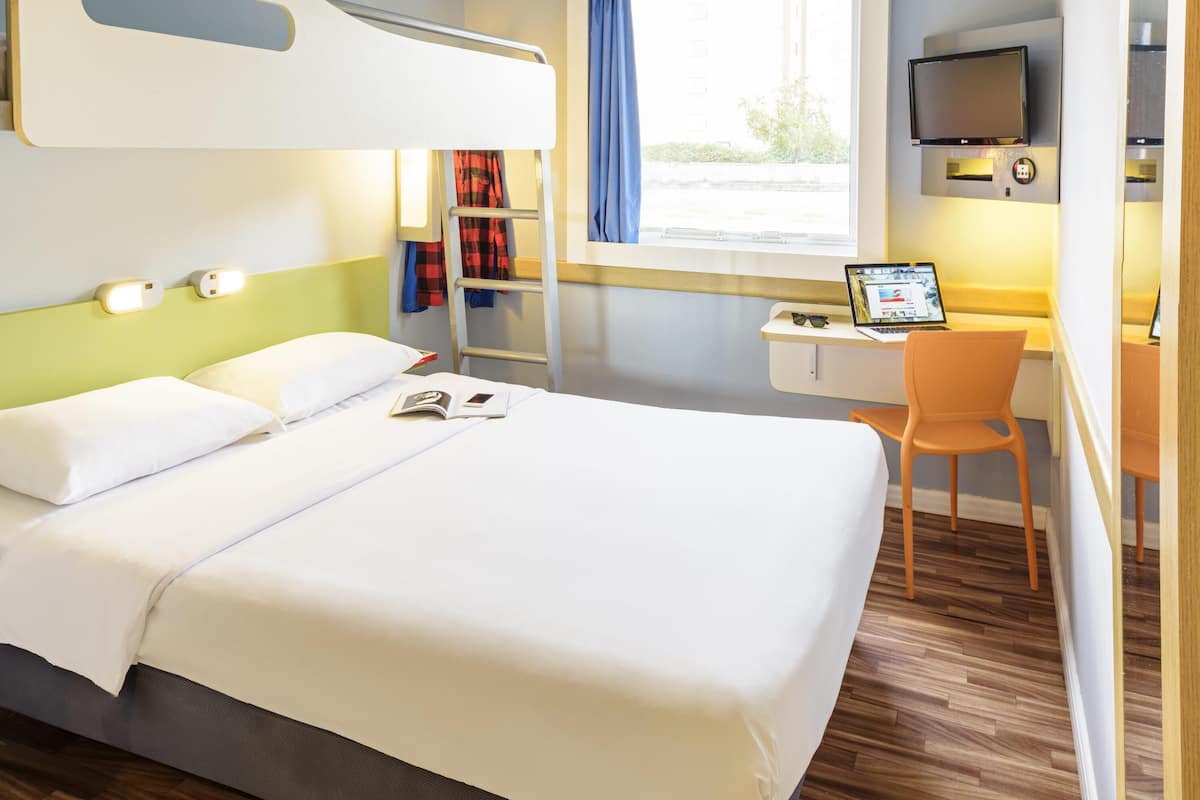 Quarto de hotel com cama, escrivaninha, cadeira, televisão e janela com cortinas.