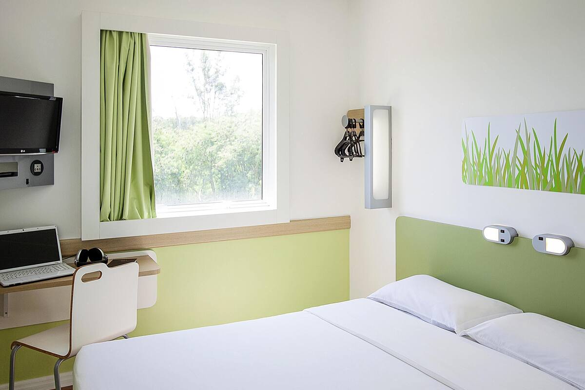 Quarto de hotel com uma cama, uma mesa com um laptop, uma televisão e uma janela com cortinas.