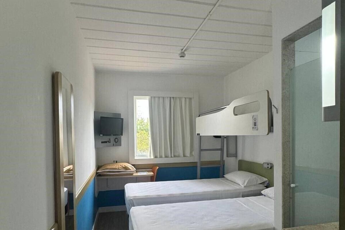 Quarto com duas camas de solteiro, uma escrivaninha com cadeira, televisão e janela com cortinas.