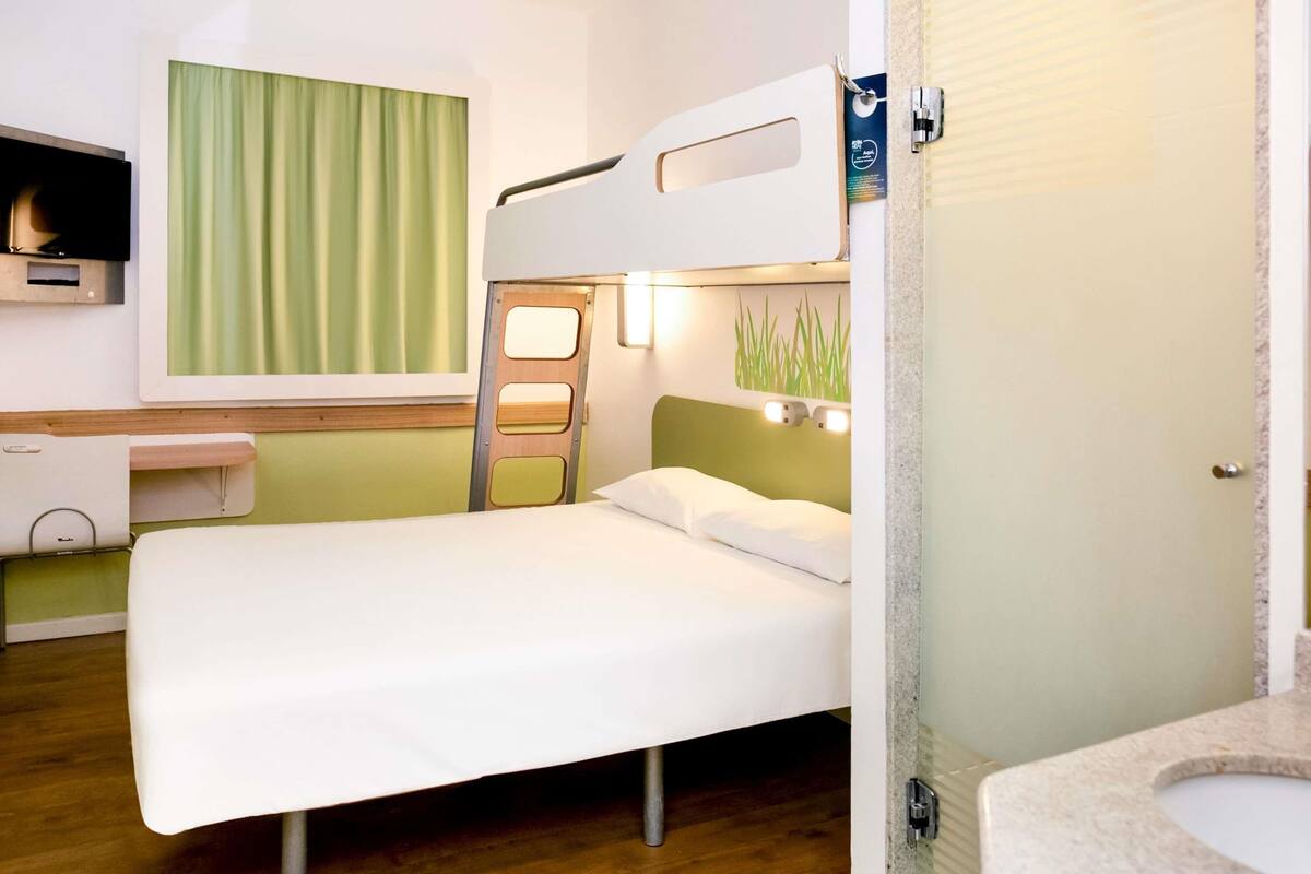 Quarto individual com uma cama beliche, uma escrivaninha, uma televisão, banheiro com pia e cortina verde.