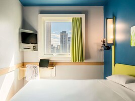 Quarto de hotel com cama, escrivaninha, laptop, televisão, janela com cortinas e vista para a cidade.