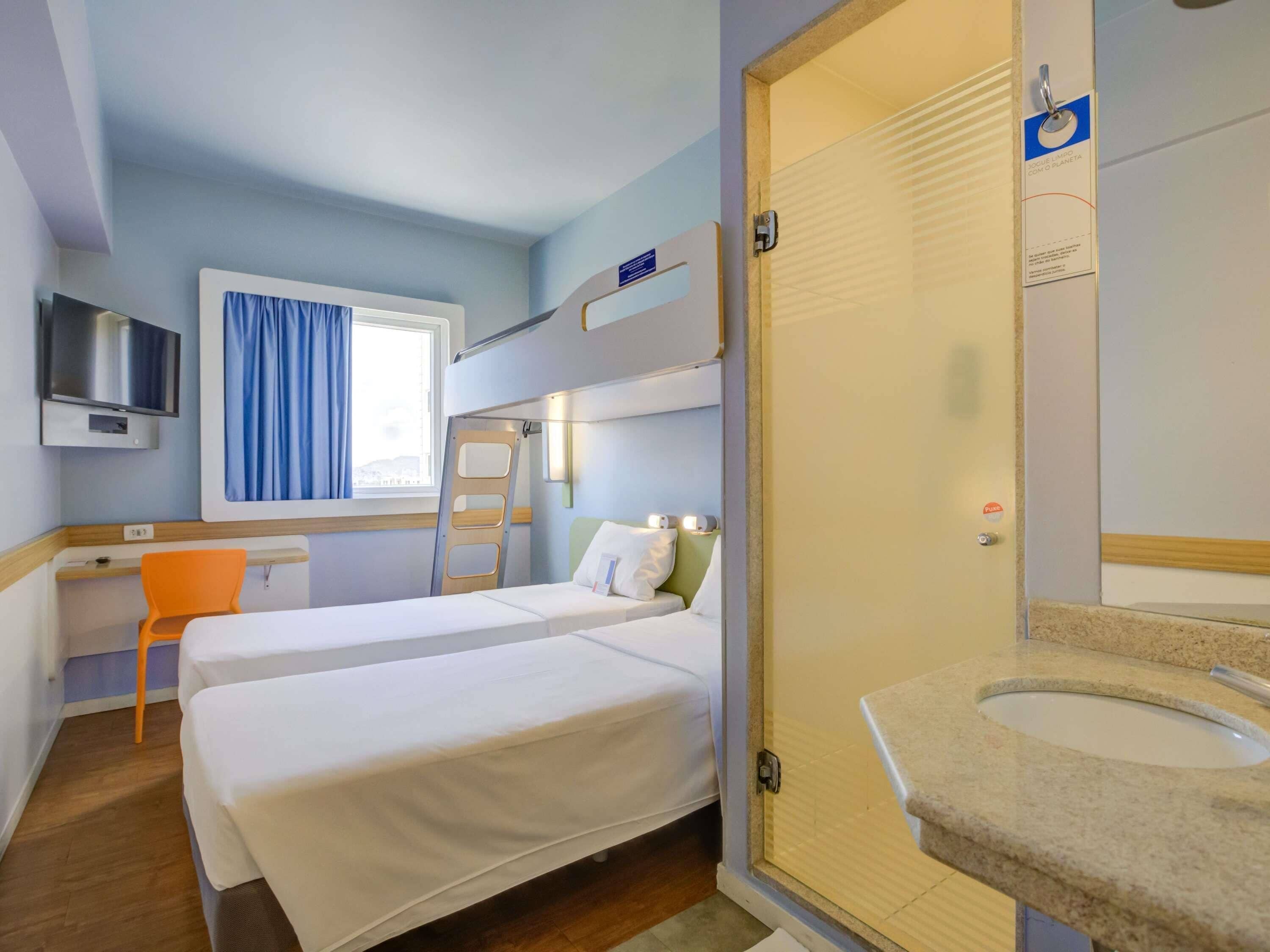 Quarto de hotel com beliches, uma escrivaninha, televisão, banheiro com pia e janela com cortinas.
