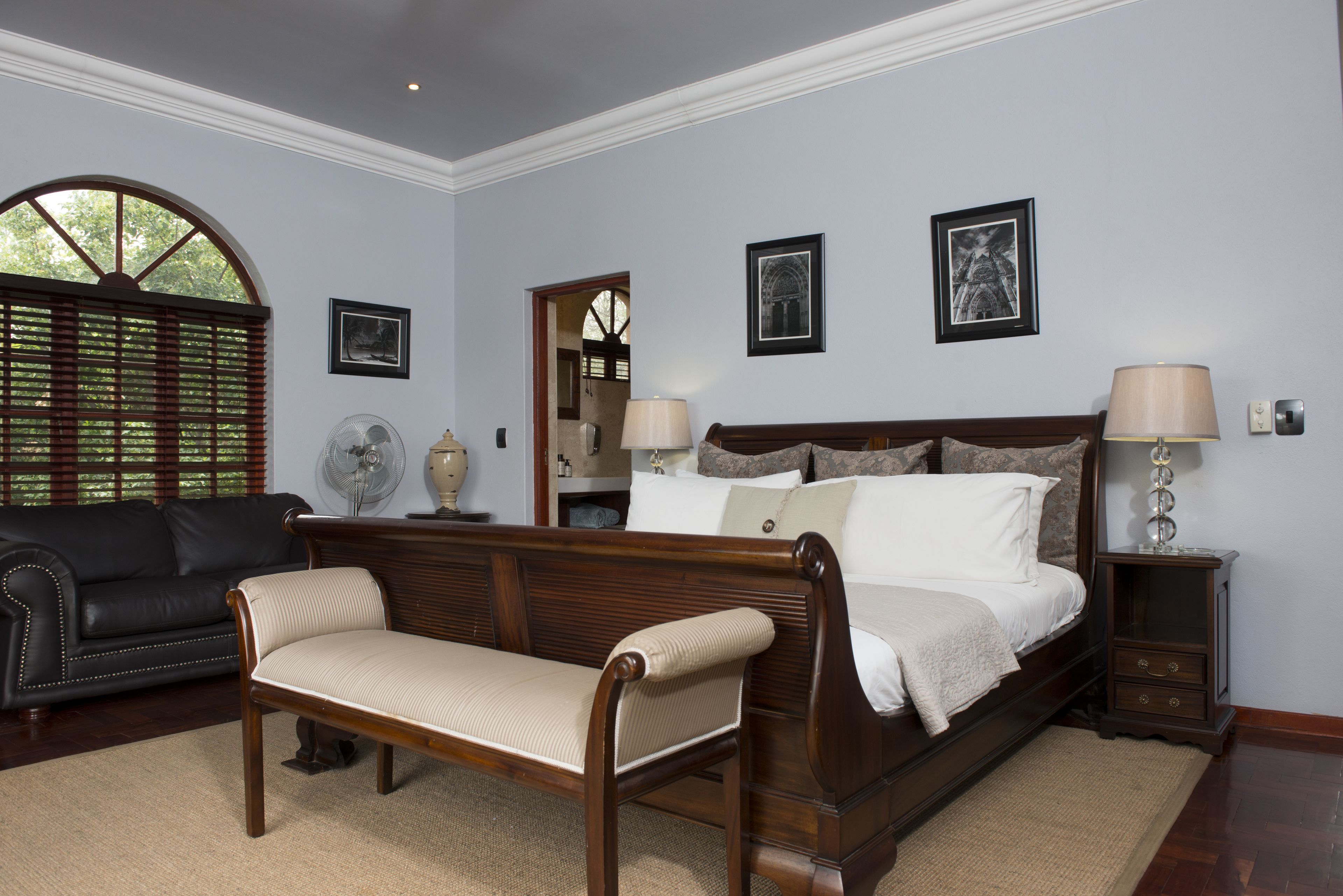 Quarto com cama de madeira, um banco, sofá de couro preto e um ventilador.
