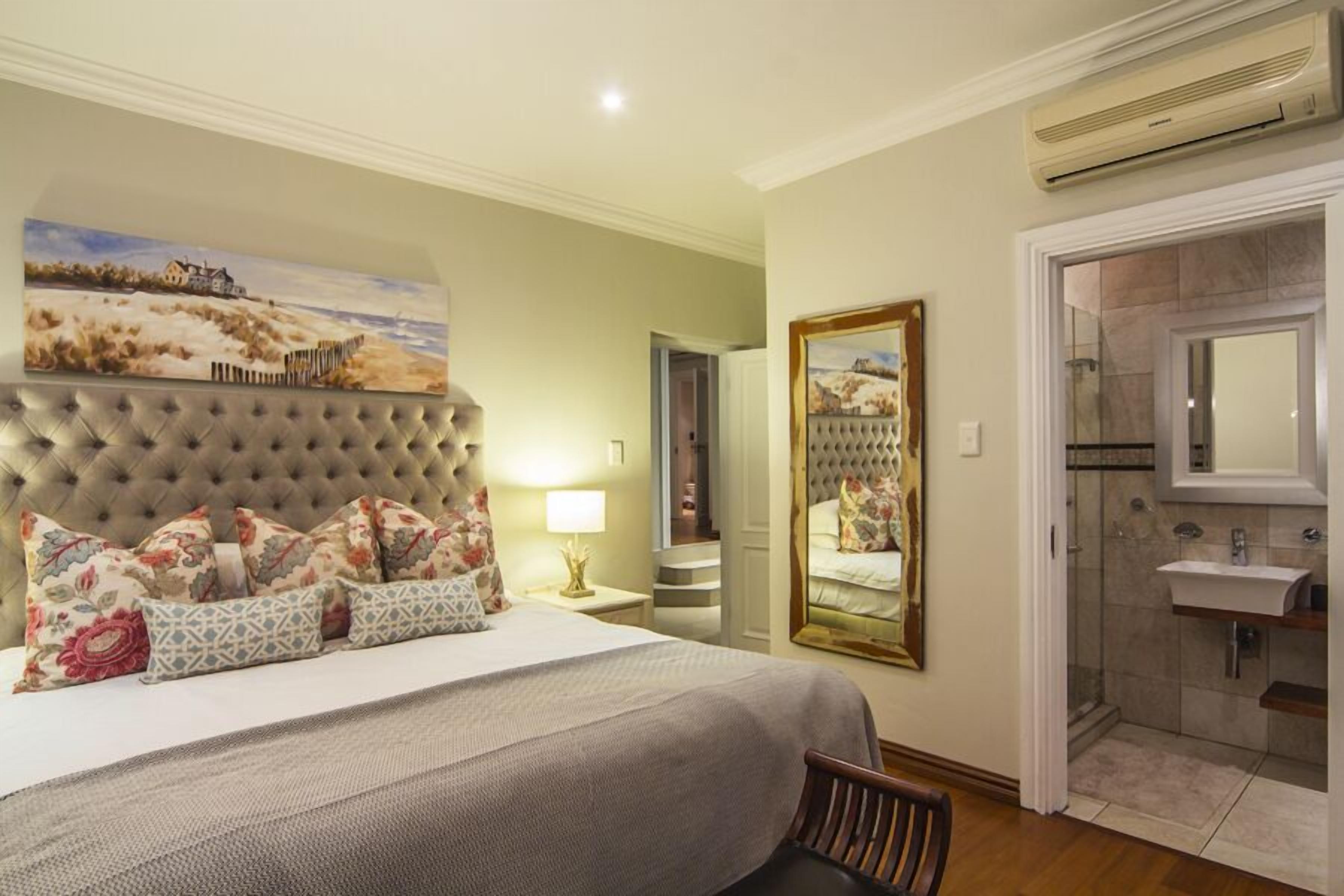 Quarto com cabeceira estofada, cama grande com almofadas estampadas, mesa de cabeceira com abajur, pintura acima da cama e banheiro com pia e espelho.