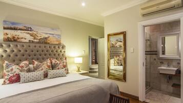Quarto com cabeceira estofada, cama grande com almofadas estampadas, mesa de cabeceira com abajur, pintura acima da cama e banheiro com pia e espelho.