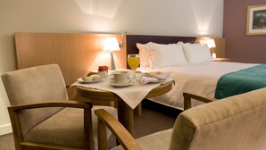 Quarto de hotel com cama, duas cadeiras, mesa com itens de café da manhã e mesa de cabeceira com luminária.