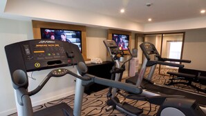 Ginásio de hotel com esteiras, um painel de controlo e duas televisões a exibir eventos desportivos.