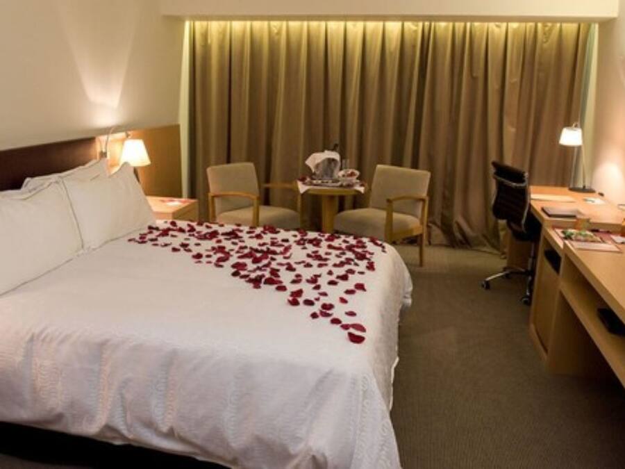 Quarto de hotel com cama, duas cadeiras, uma mesinha, escrivaninha e televisão.