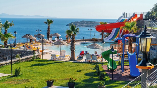 Área de piscina em resort com toboáguas, espreguiçadeiras, guarda-sóis e vista para o mar.