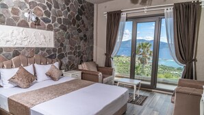 Quarto com parede de pedra, uma cama grande, duas cadeiras, uma mesa de cabeceira e vista para o oceano.