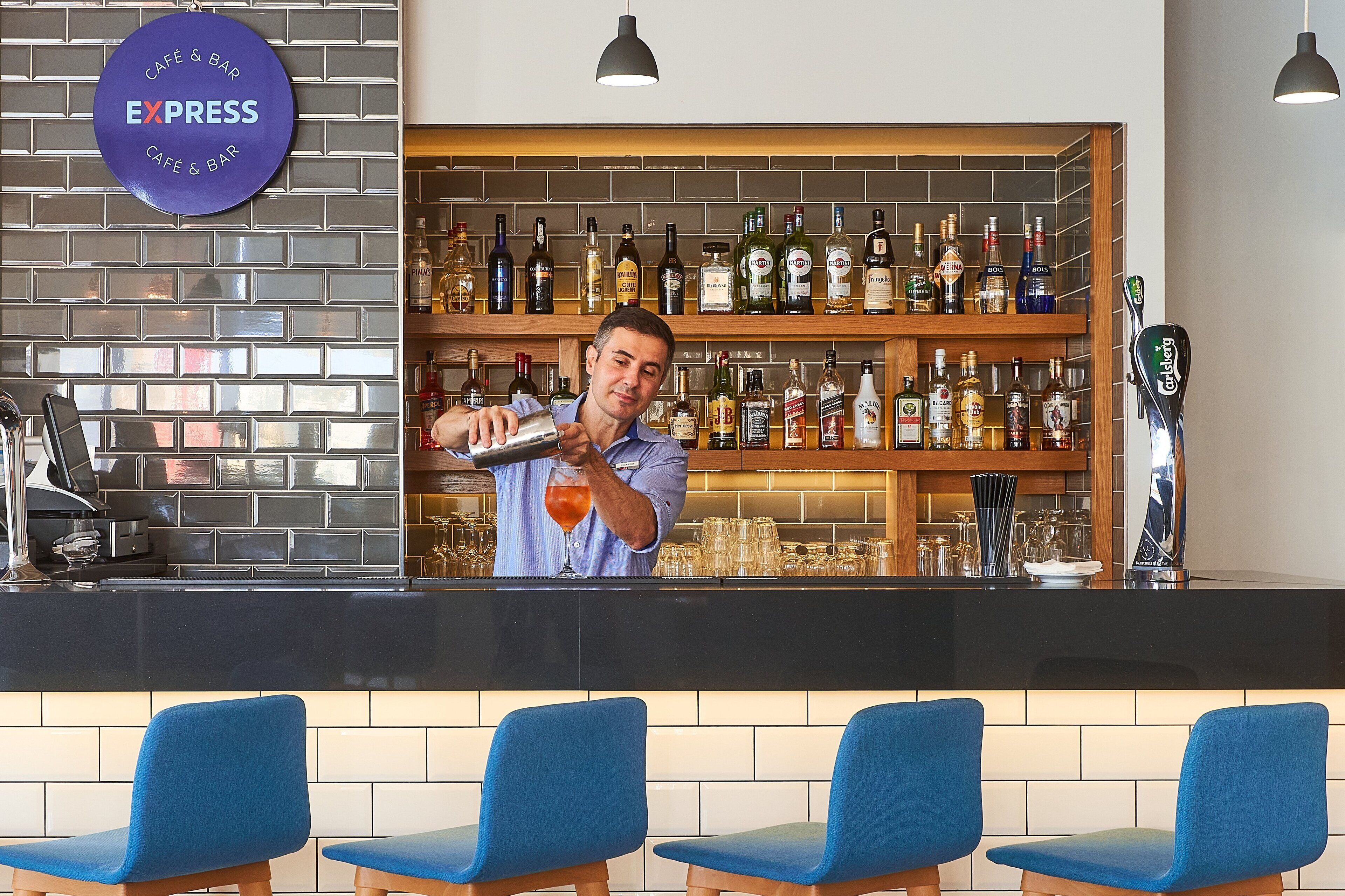 Uma área de bar com um bartender preparando uma bebida, prateleiras com garrafas de bebidas e cadeiras azuis.