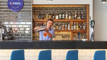 Uma área de bar com um bartender preparando uma bebida, prateleiras com garrafas de bebidas e cadeiras azuis.