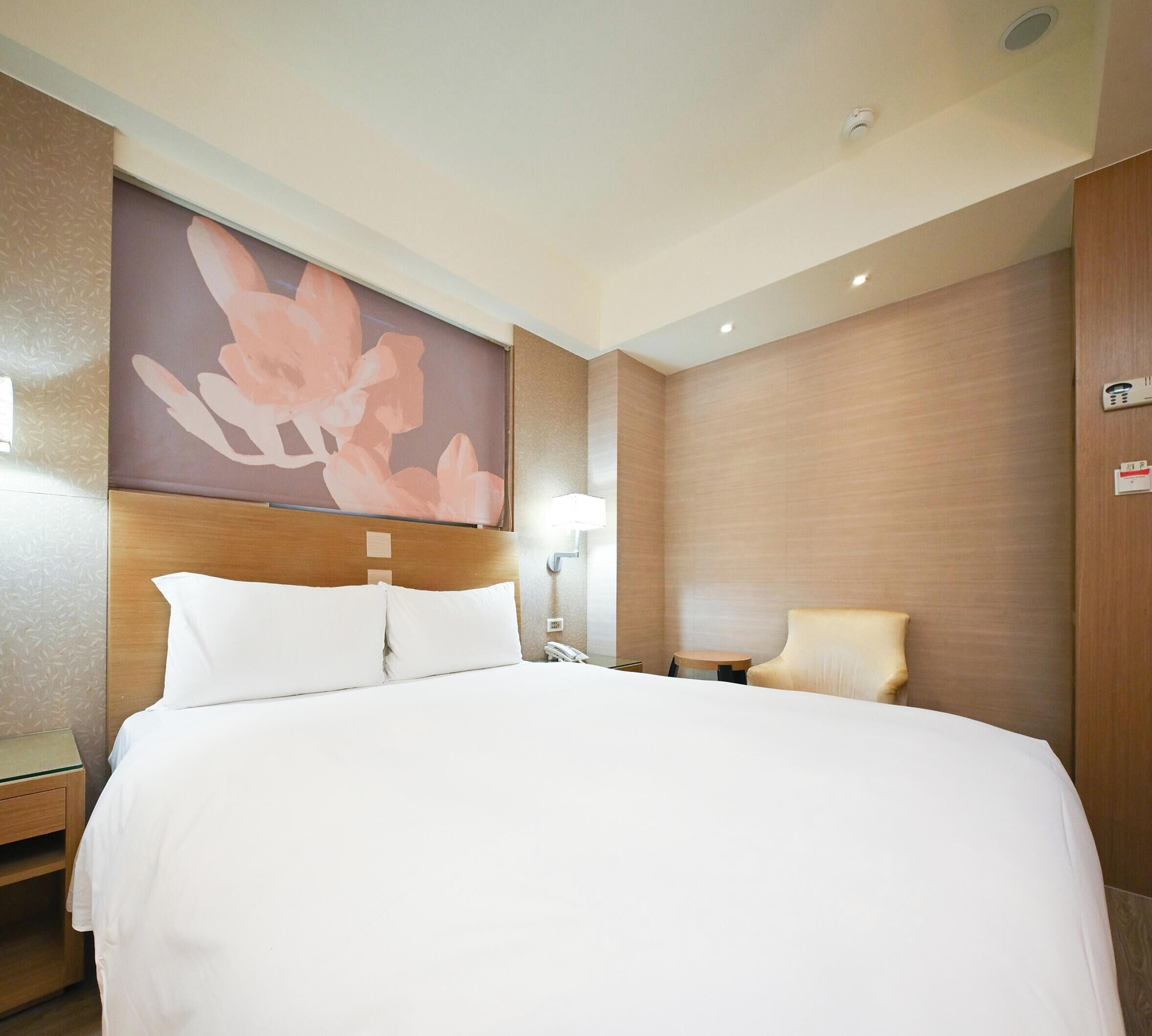Quarto de hotel com uma cama grande, uma mesa de cabeceira, uma cadeira e uma luminária fixada na parede.