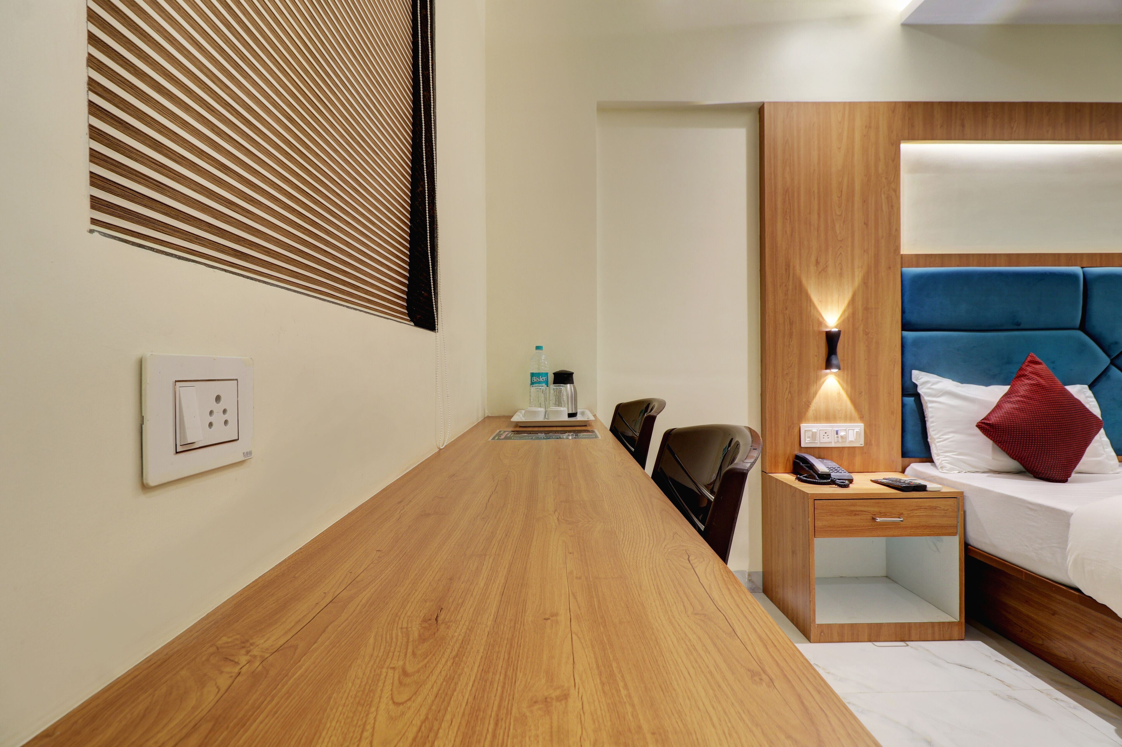 Quarto de hotel com uma mesa de jantar de madeira, uma cama com cabeceira azul, uma mesa de cabeceira com luminária e uma tomada na parede.