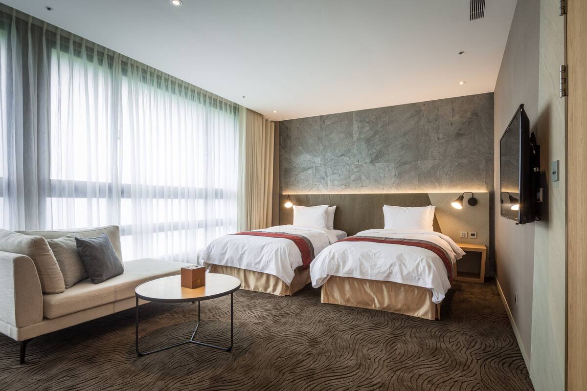 Quarto de hotel com duas camas, um sofá, uma mesa pequena e uma TV fixada na parede.
