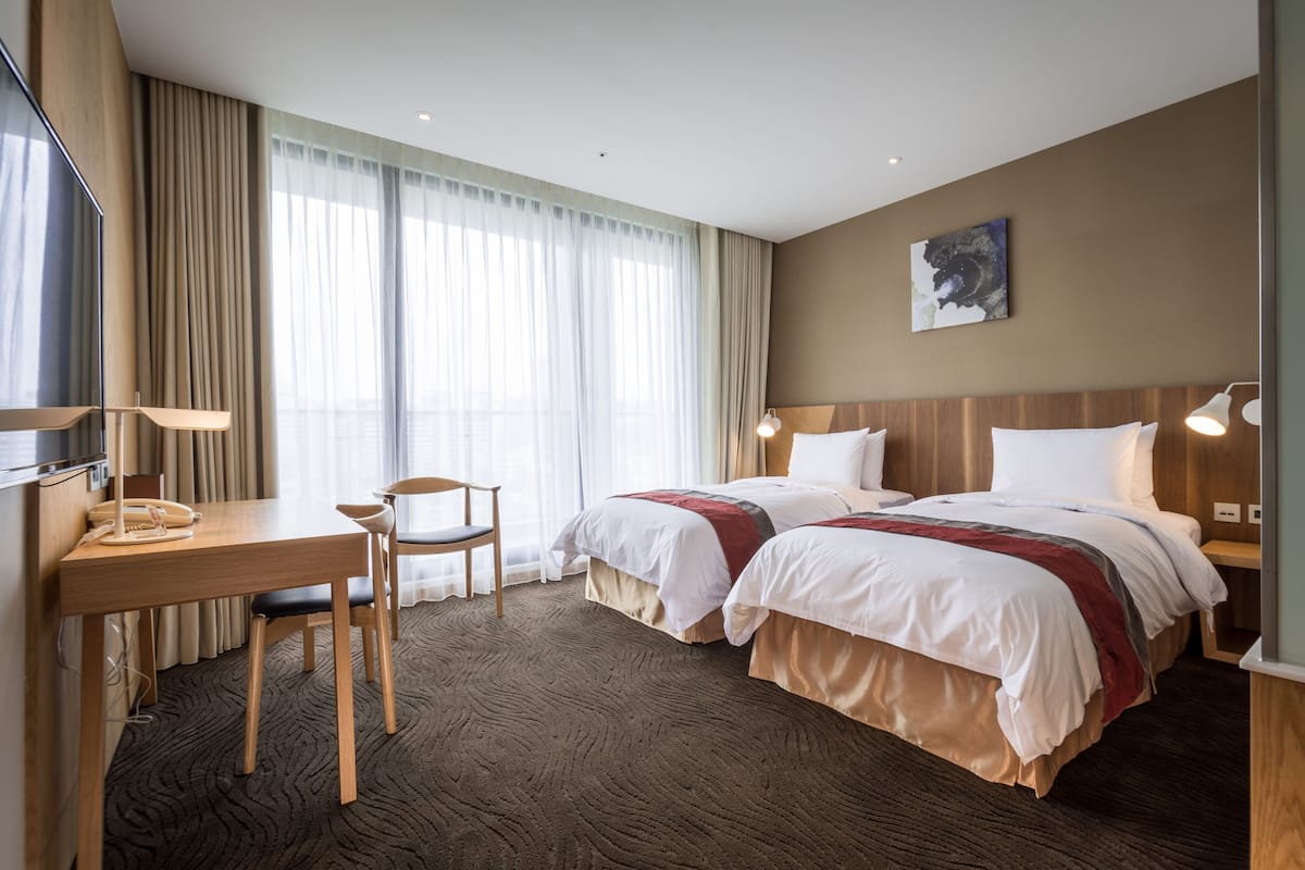 Quarto de hotel com duas camas, uma escrivaninha com cadeira, uma janela grande com cortinas e um quadro na parede.