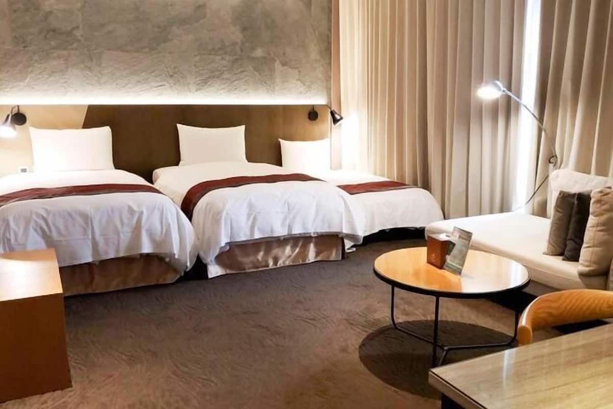Quarto de hotel com duas camas, um sofá, uma mesa pequena e um abajur.