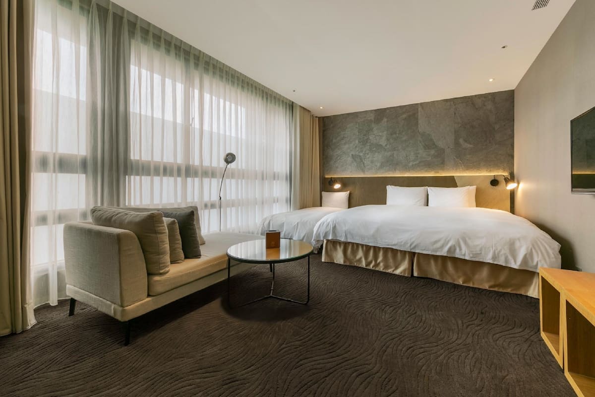 Quarto de hotel moderno com uma cama grande, um sofá, uma mesinha redonda e uma janela ampla com cortinas leves.