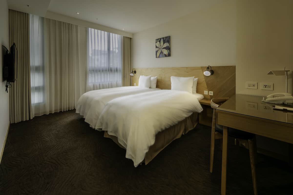 Quarto de hotel com uma cama grande, uma escrivaninha, uma cadeira, uma televisão e uma janela com cortinas.
