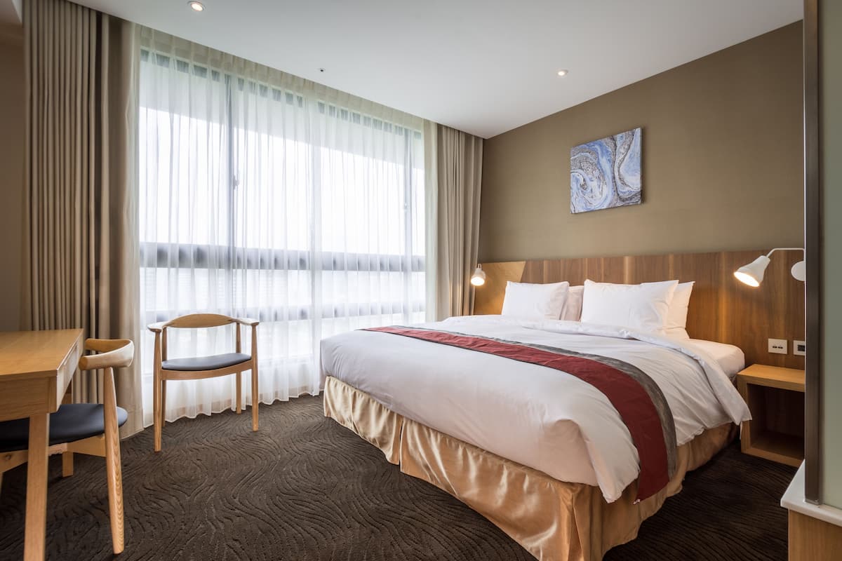 Quarto de hotel com uma cama grande, uma escrivaninha, uma cadeira e uma janela com cortinas translúcidas.