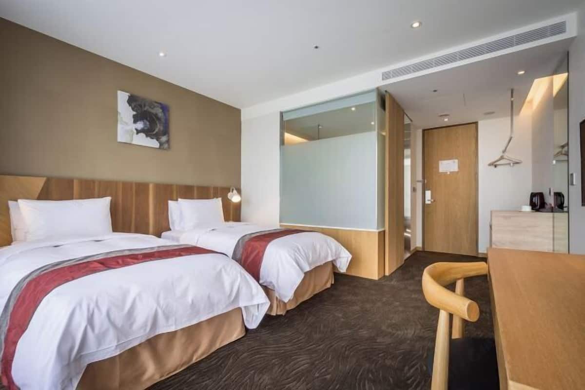 Quarto de hotel com duas camas, uma escrivaninha, uma cadeira, um porta-roupas e um espelho.