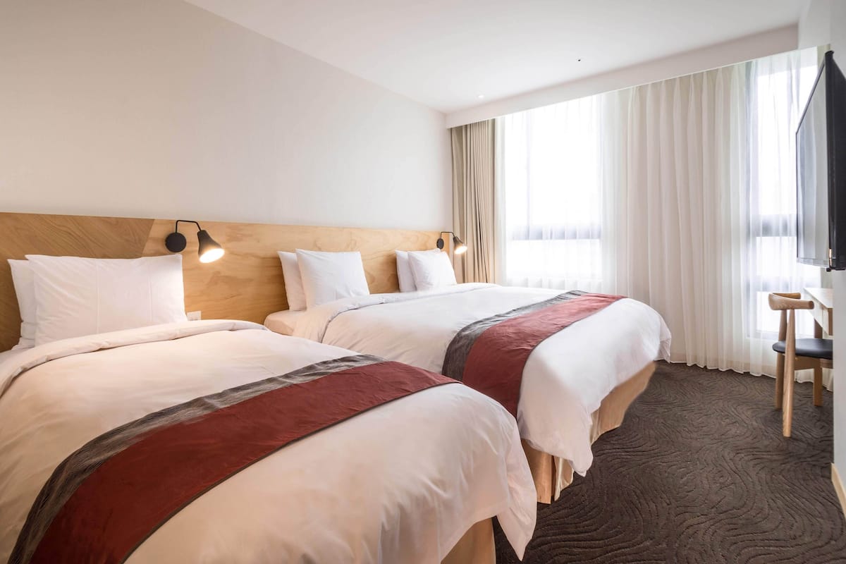 Quarto de hotel com duas camas, cabeceira de madeira, uma mesinha e uma cadeira.