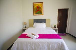Uma cama bem arrumada com lençóis brancos e um toque de rosa, uma luminária de cabeceira, um quadro emoldurado e uma porta de madeira.