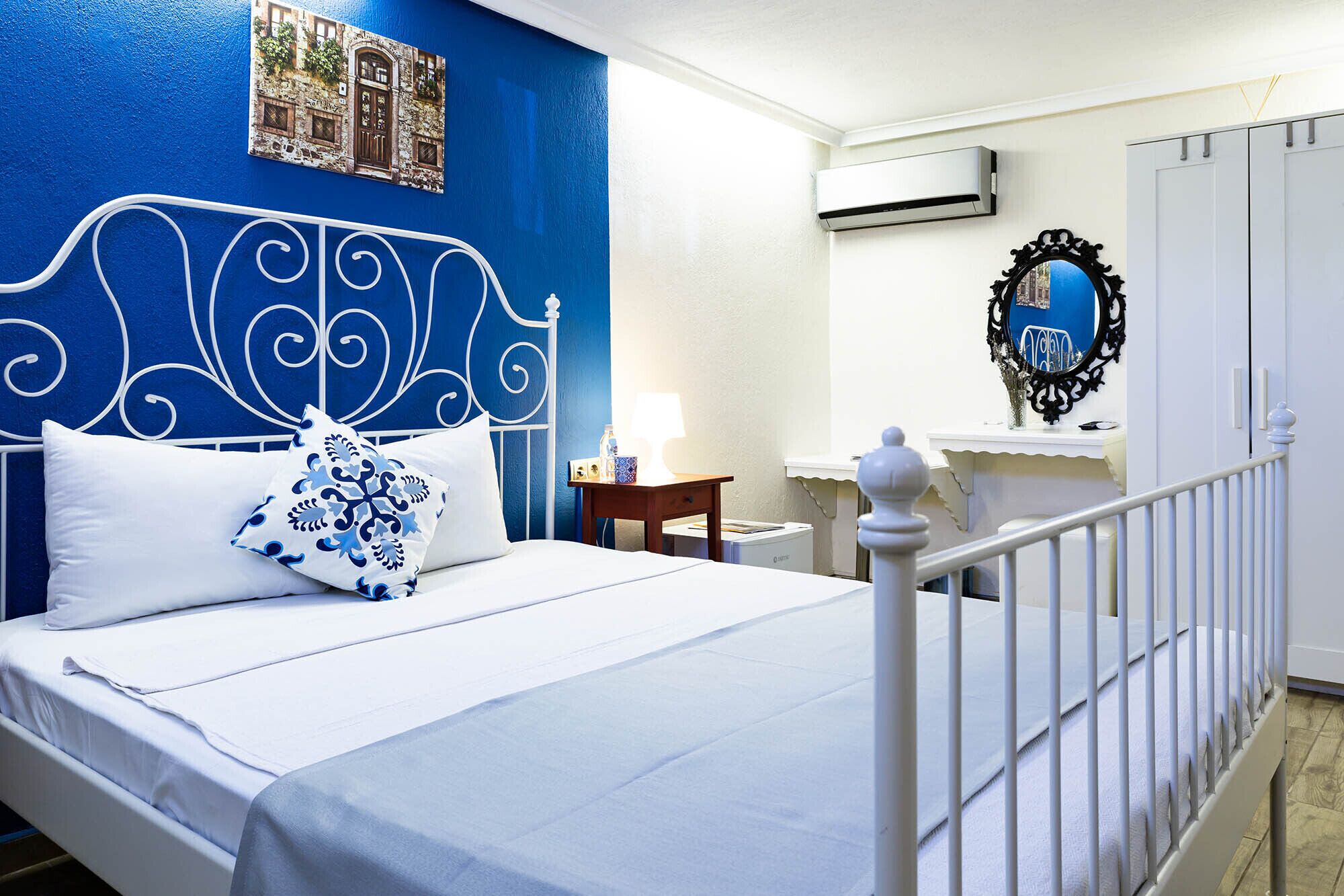 Quarto com cama branca, paredes azuis, cabeceira decorativa, mesa de cabeceira pequena com abajur e um guarda-roupa.