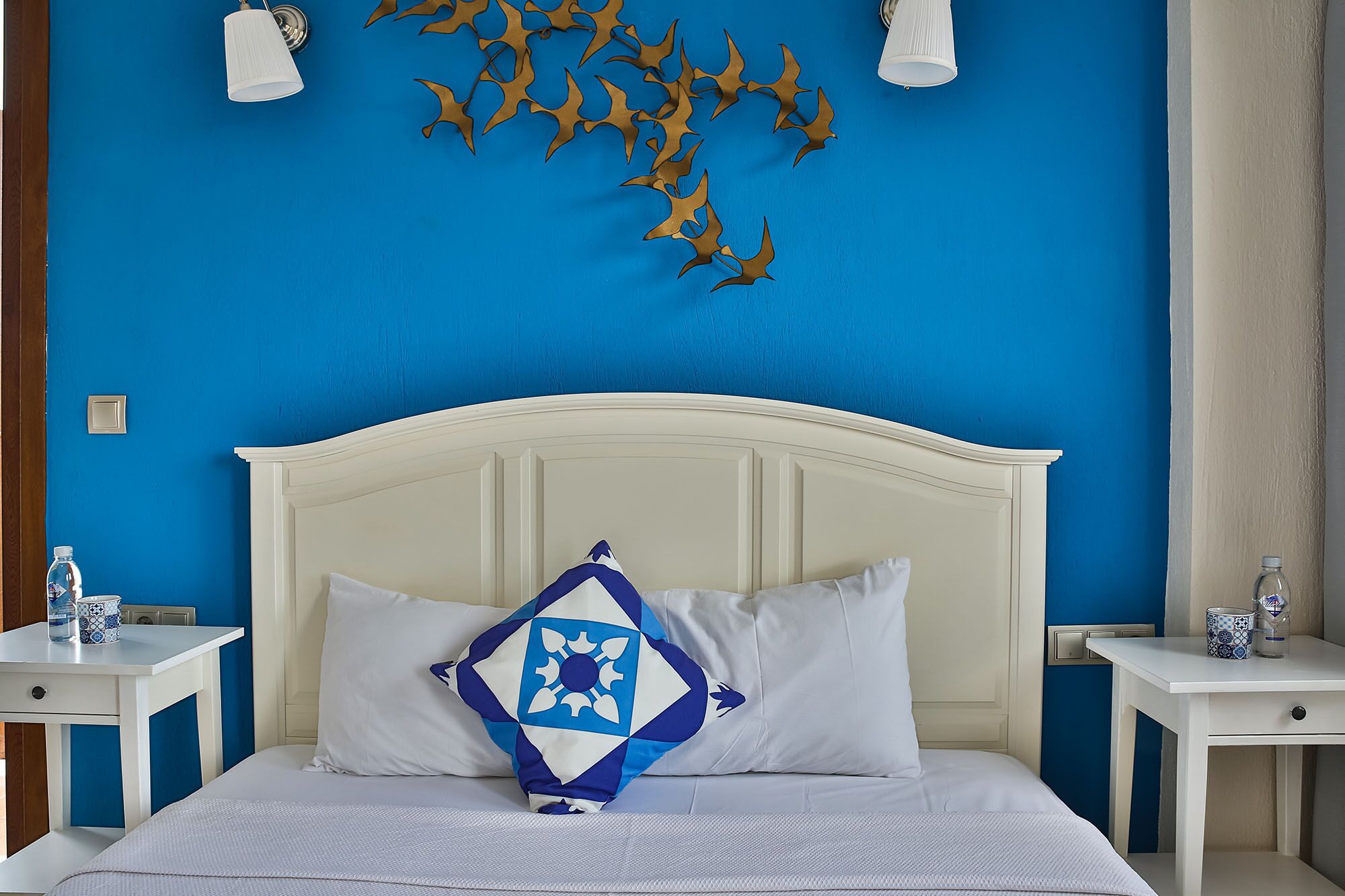 Quarto com cabeceira branca, almofada estampada em azul e branco, e decoração de pássaros dourados na parede.