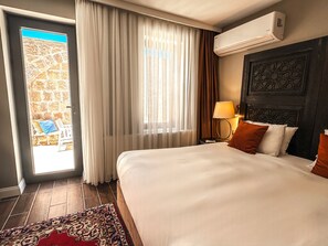 Quarto de hotel com cama, mesa de cabeceira, abajur, ar condicionado e vista através de uma porta.