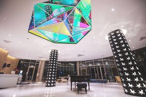 Um lobby moderno com uma instalação de iluminação geométrica, colunas pretas com desenhos de cavalos, um piano de cauda e um balcão de recepção.