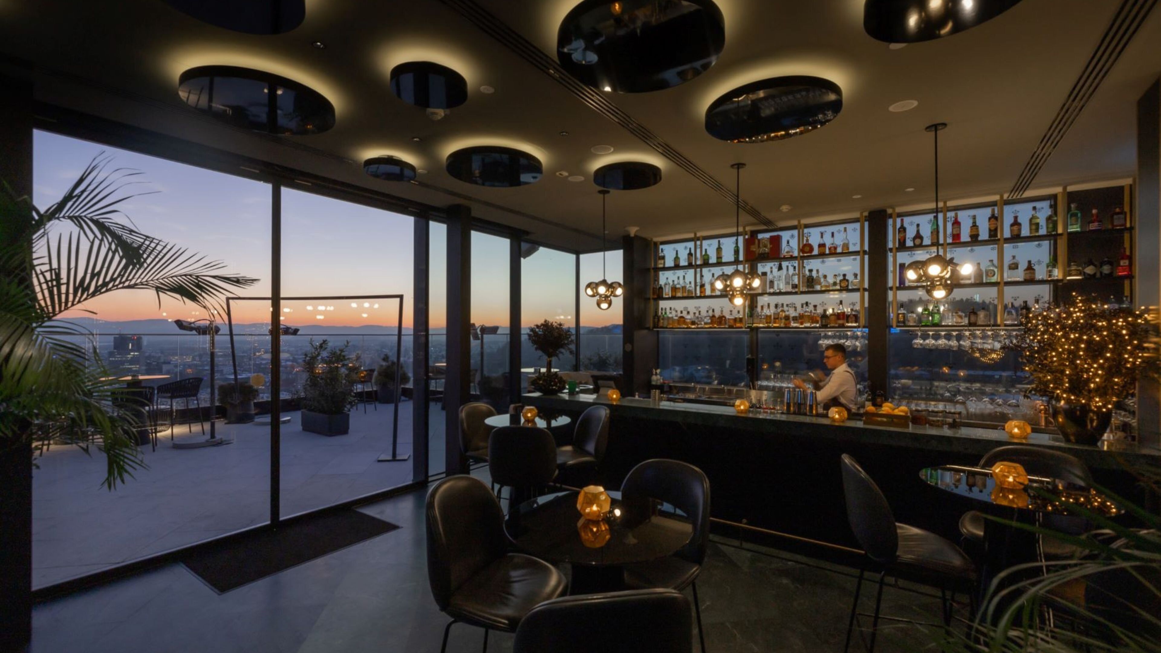 Uma área de bar com um bartender, grandes janelas e vista para a cidade.