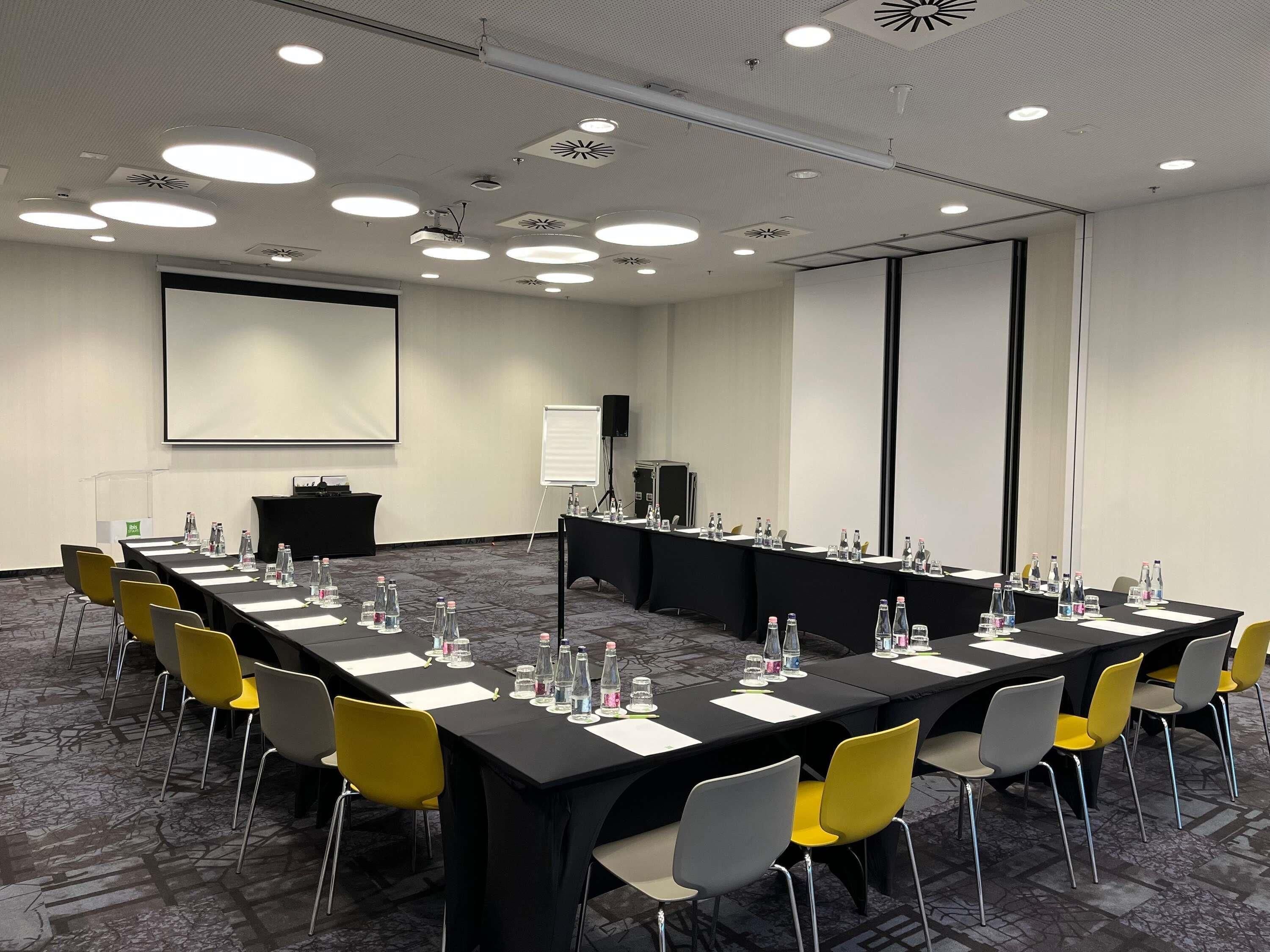 Uma sala de conferência com uma mesa comprida, cadeiras, uma tela de projetor e um quadro branco.
