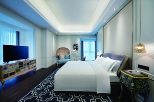 Quarto de hotel moderno com uma cama grande, televisão, área de estar com uma poltrona e sofá, e uma mesinha decorativa.