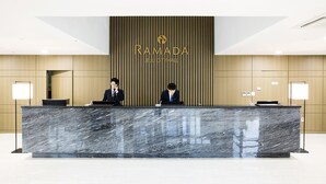 Uma receção com bancada de mármore, duas recepcionistas, e um letreiro acima que diz "RAMADA JEJU CITYHALL".