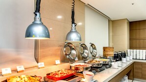 Disposição de buffet com diversos alimentos, incluindo uma tigela vermelha de frango frito, uma tigela branca de sopa e vários recipientes de metal com comida.