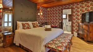Quarto com uma cama grande, cabeceira de madeira, um banco, mesa de cabeceira, cômoda, televisão e banheiro visível por uma porta aberta.