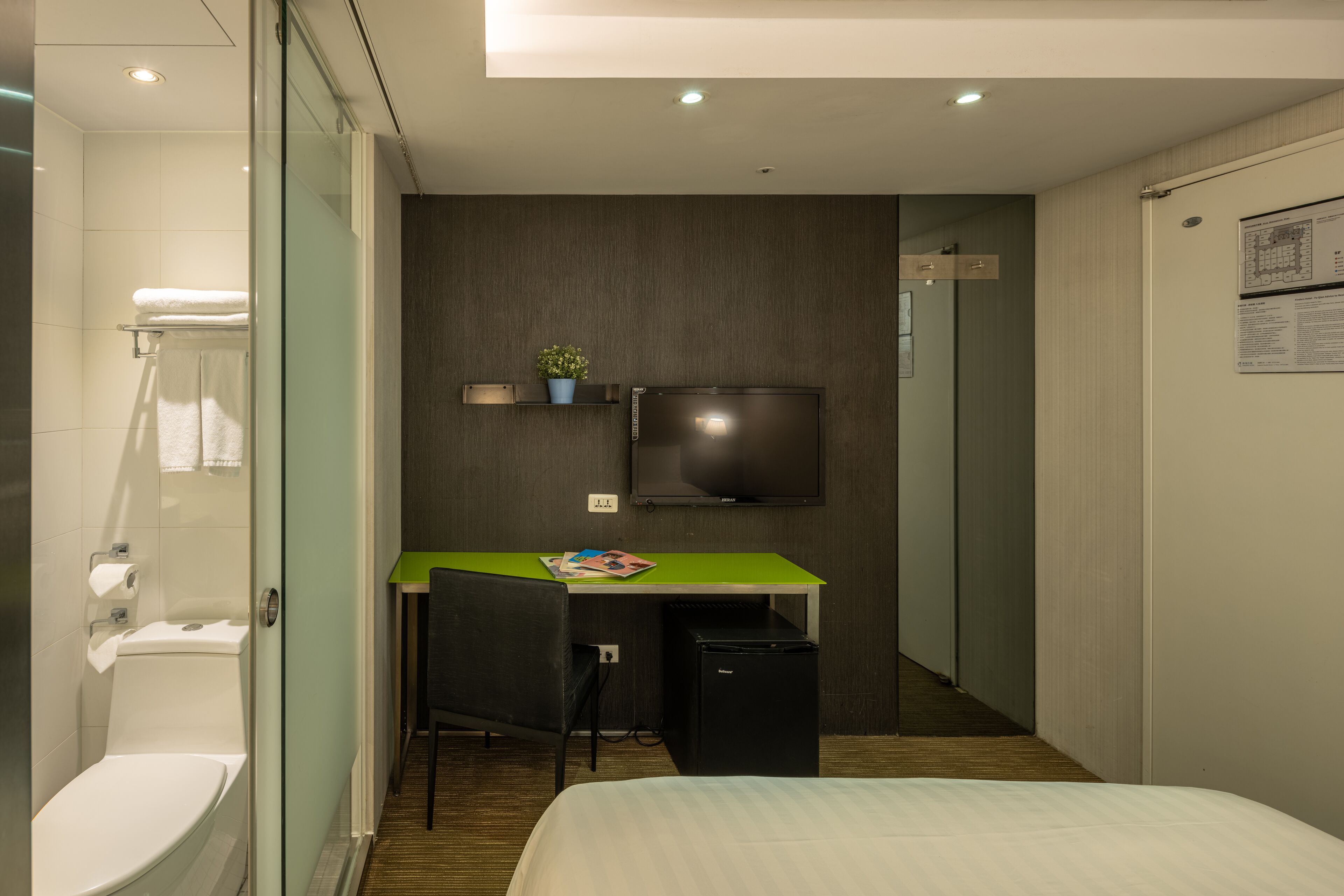 Quarto de hotel com uma cama, uma mesa com cadeira, uma pequena lixeira, televisão na parede e área de chuveiro.