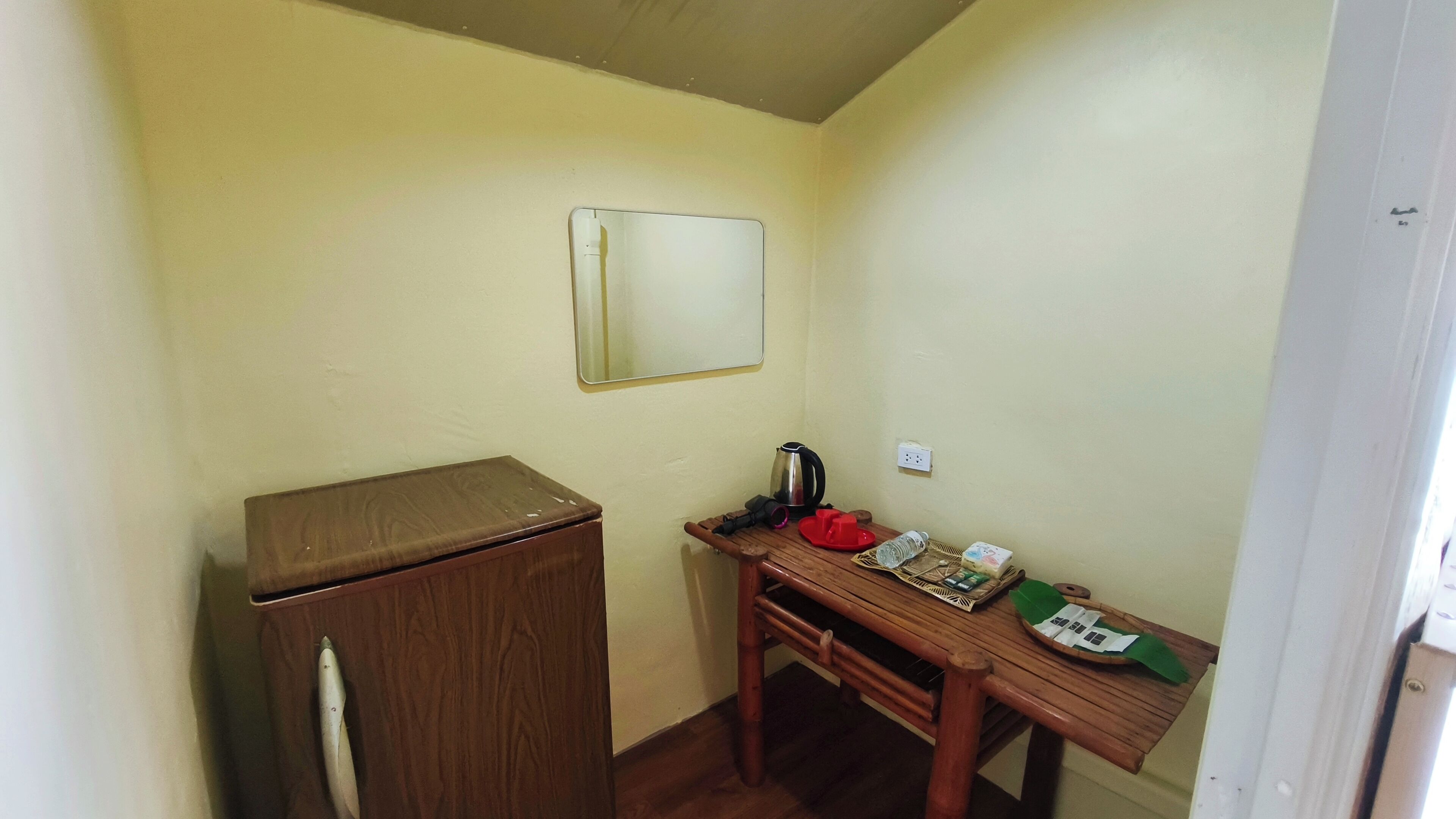 Quarto compacto com um armário de madeira, uma mesa pequena com chaleira, um tabuleiro vermelho e um espelho na parede.