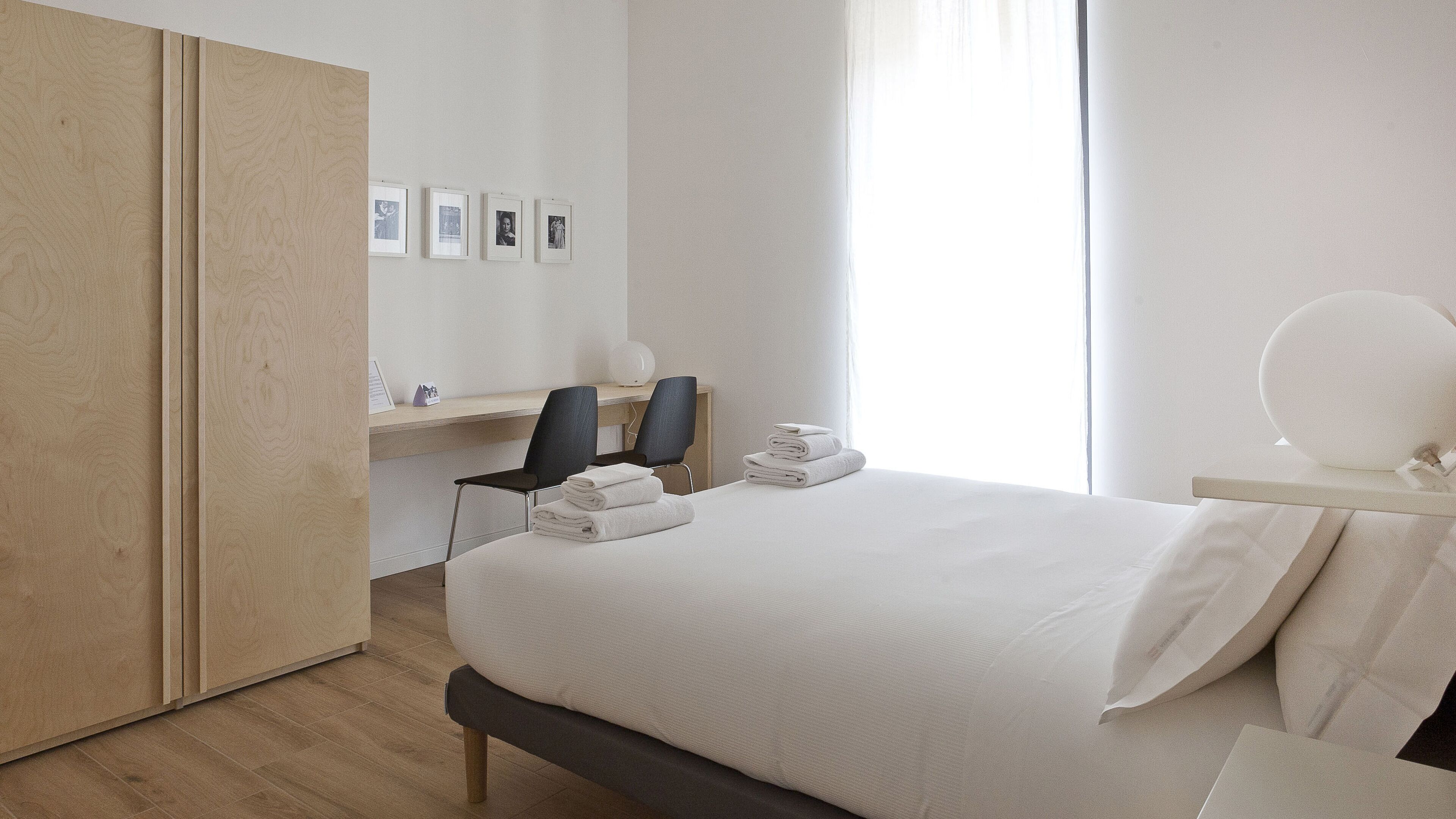 Quarto de hotel com cama, escrivaninha, cadeira, guarda-roupa e mesa de cabeceira com abajur.