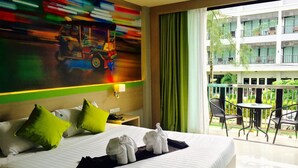 Quarto de hotel com uma cama grande, almofadas verdes, um quadro na parede, uma varanda com cadeiras e uma mesa, e vista para um edifício.