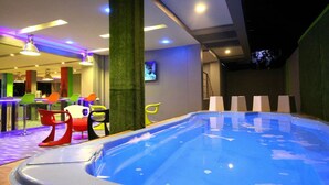 Piscina com cadeiras coloridas, área de bar e uma tela de televisão.