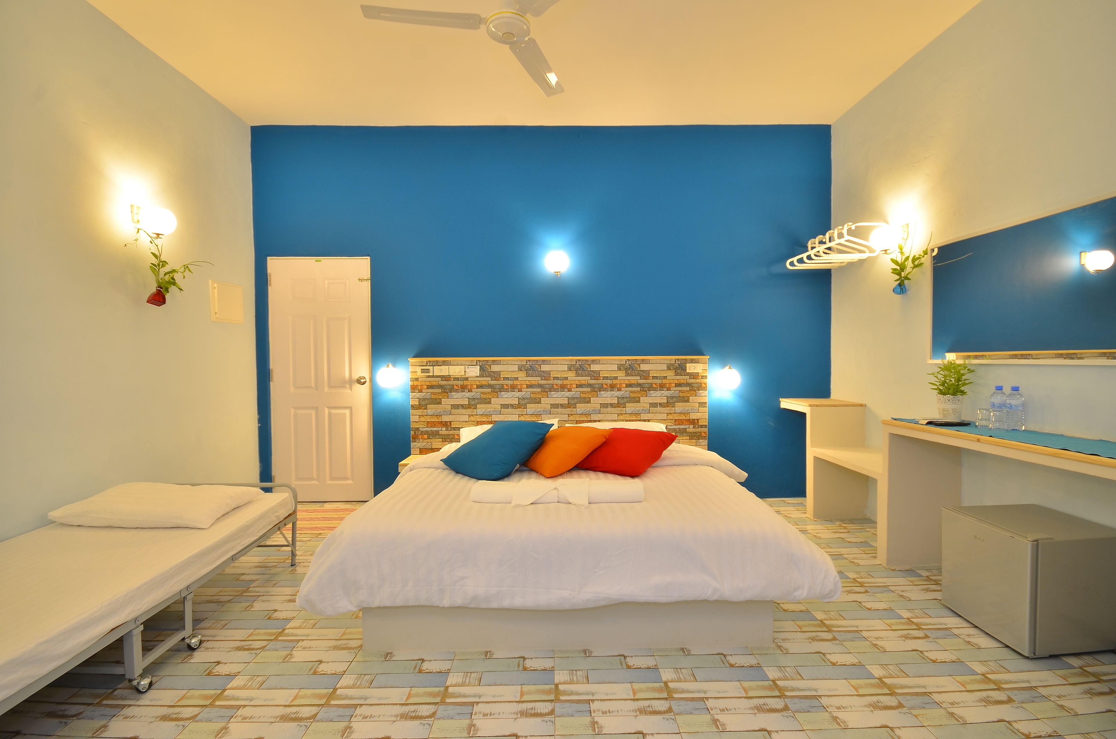 Quarto com cama, mesas de cabeceira, escrivaninha, cadeira, ventilador de teto e porta.