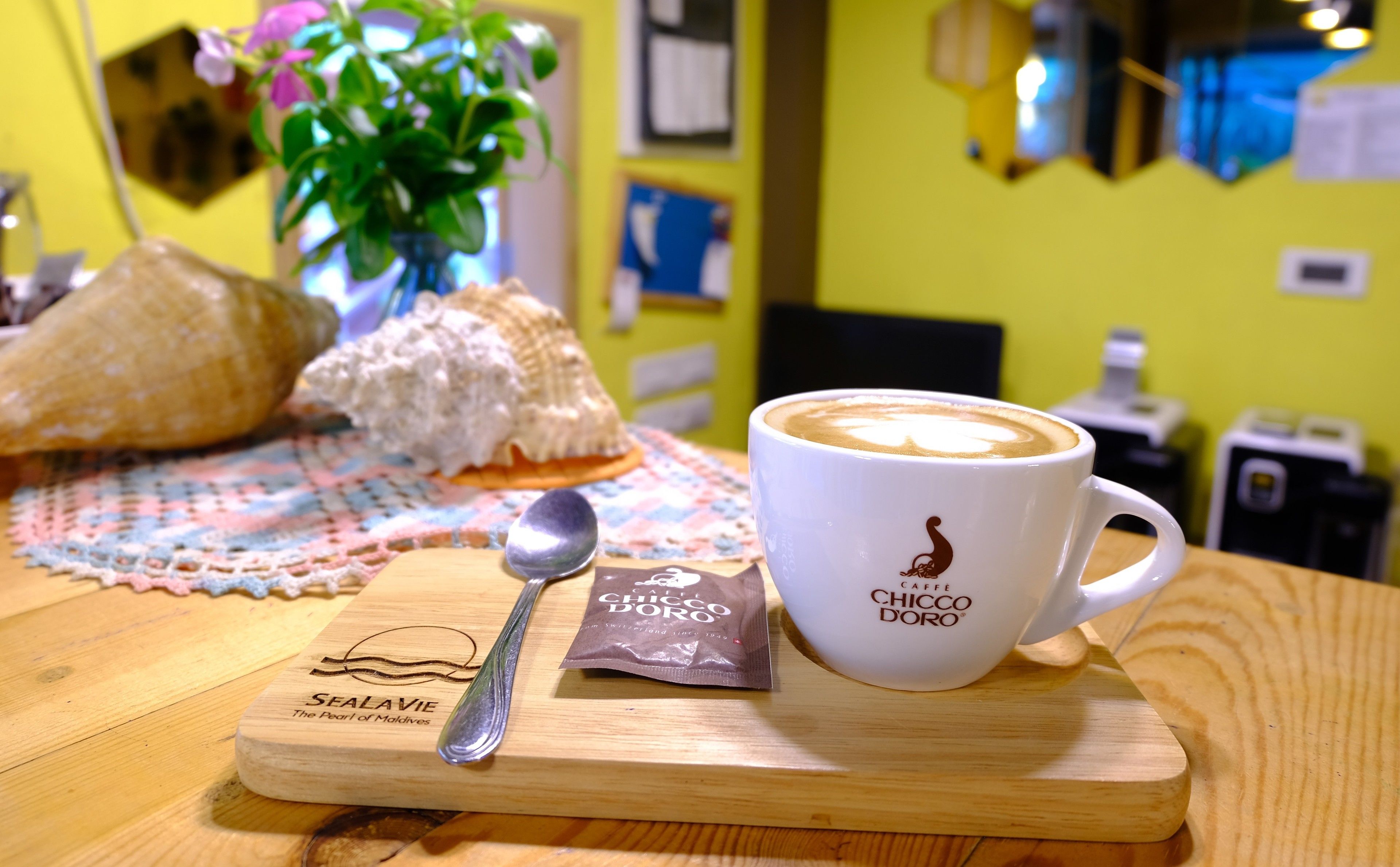 Uma chávena de café com arte de latte sobre uma mesa de madeira, uma colher, um saquinho de biscotti e uma concha decorativa.