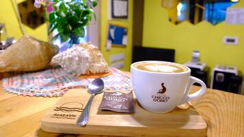 Uma chávena de café com arte de latte sobre uma mesa de madeira, uma colher, um saquinho de biscotti e uma concha decorativa.