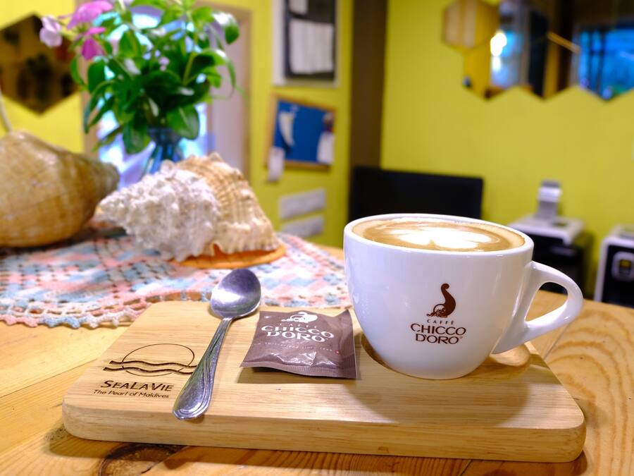 Uma chávena de café com arte de latte sobre uma mesa de madeira, uma colher, um saquinho de biscotti e uma concha decorativa.