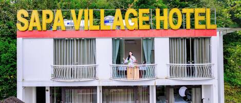 Um edifício de dois andares com a placa 'SAPA VILLAGE HOTEL' sobre a entrada. Possui varandas com grades e uma pessoa parada em uma delas.