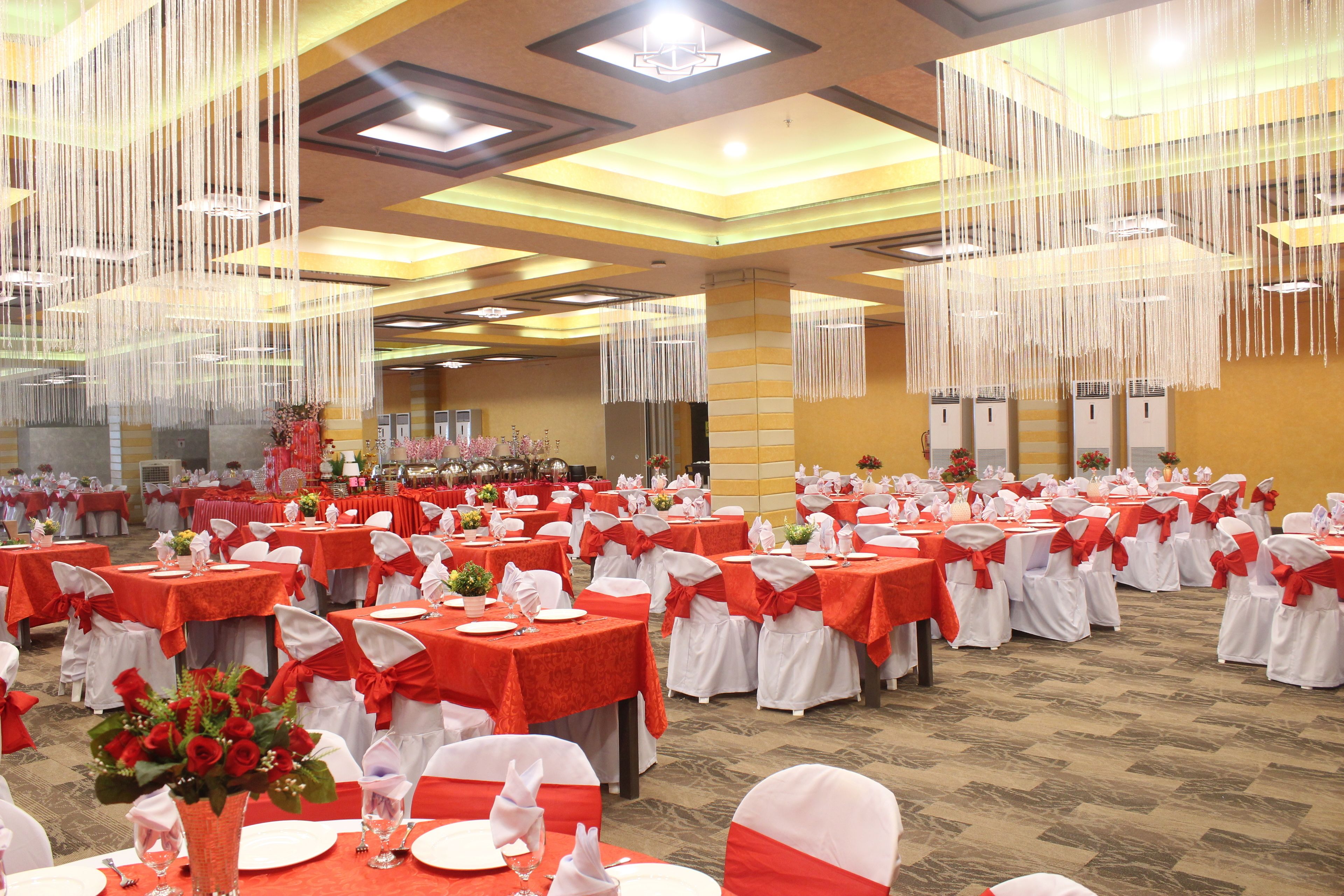 Uma sala de banquetes com várias mesas redondas, cada uma com pratos brancos, toalhas vermelhas e cadeiras brancas adornadas com fitas vermelhas.