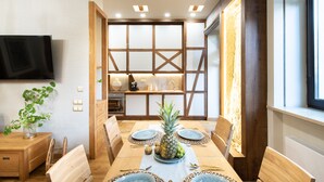 Uma sala de jantar com uma mesa de madeira posta para dois, um arranjo de abacaxi no centro e uma TV montada na parede.