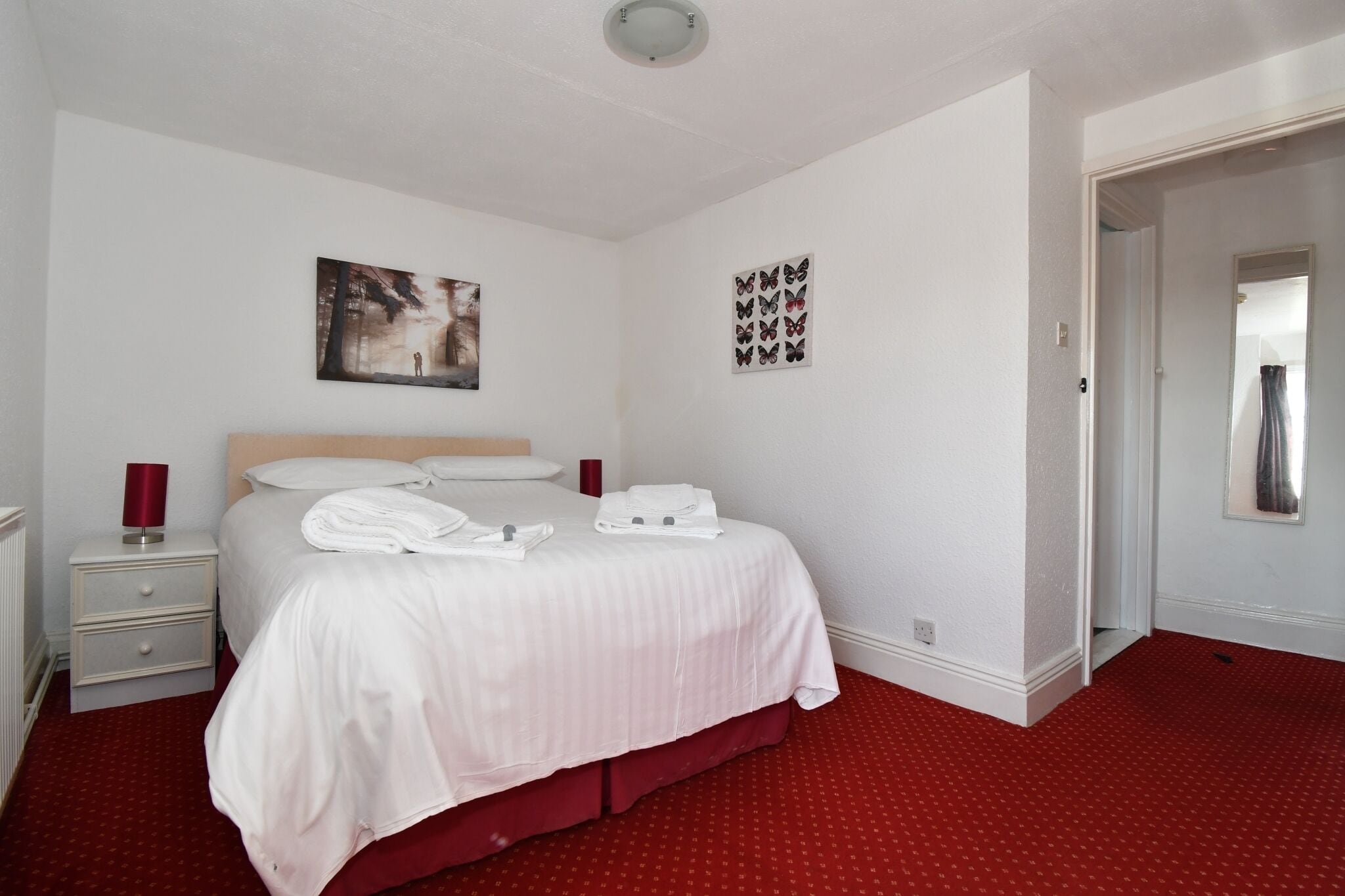 Uma cama bem arrumada com roupa de cama branca, uma pequena mesa de cabeceira com abajur vermelho e um quadro fixado na parede.